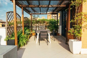 Um alpendre coberto com mesa e cadeiras, rodeado por plantas em vasos e um cercado de madeira.