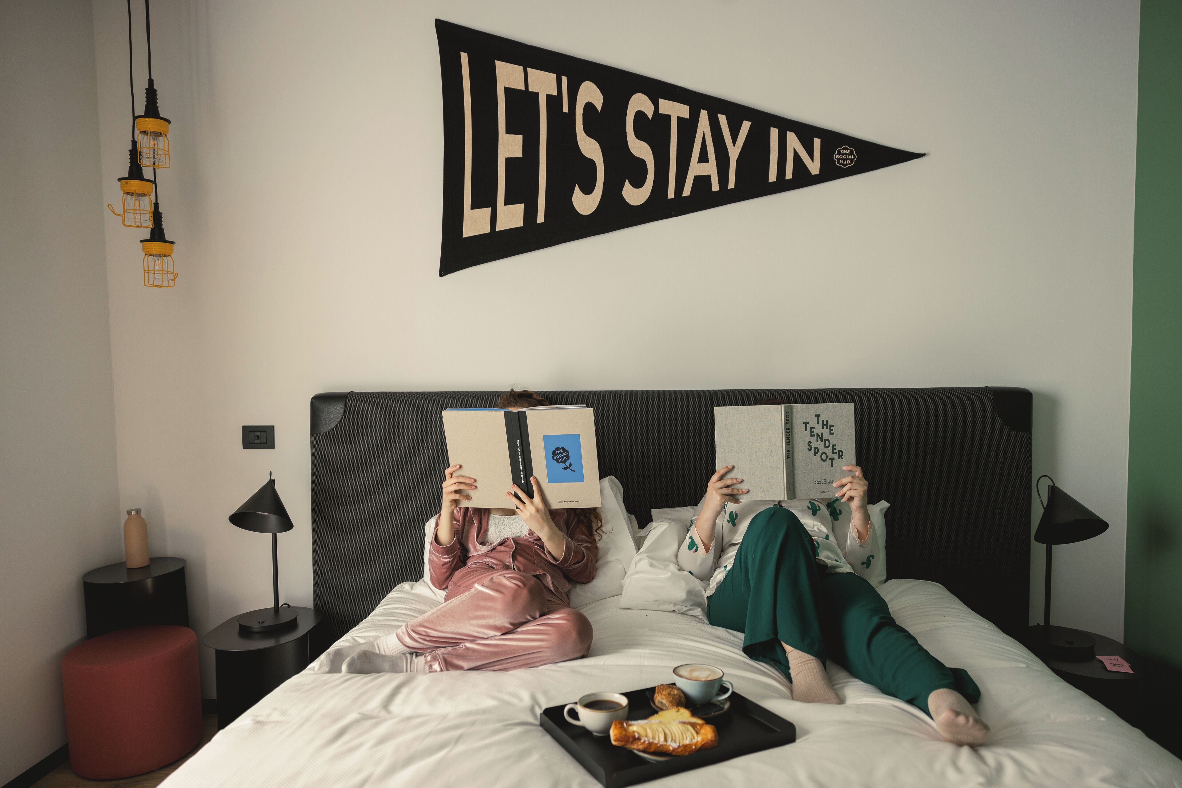 Duas pessoas leem livros num quarto com cama, mesas de cabeceira e um letreiro na parede.
