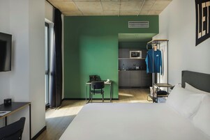 Quarto de hotel com cama, escrivaninha, cadeira, cozinha compacta e televisão na parede.