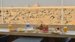 Mesa de café da manhã com diversos alimentos e uma garrafa de água, com vista para uma paisagem desértica com pirâmides.