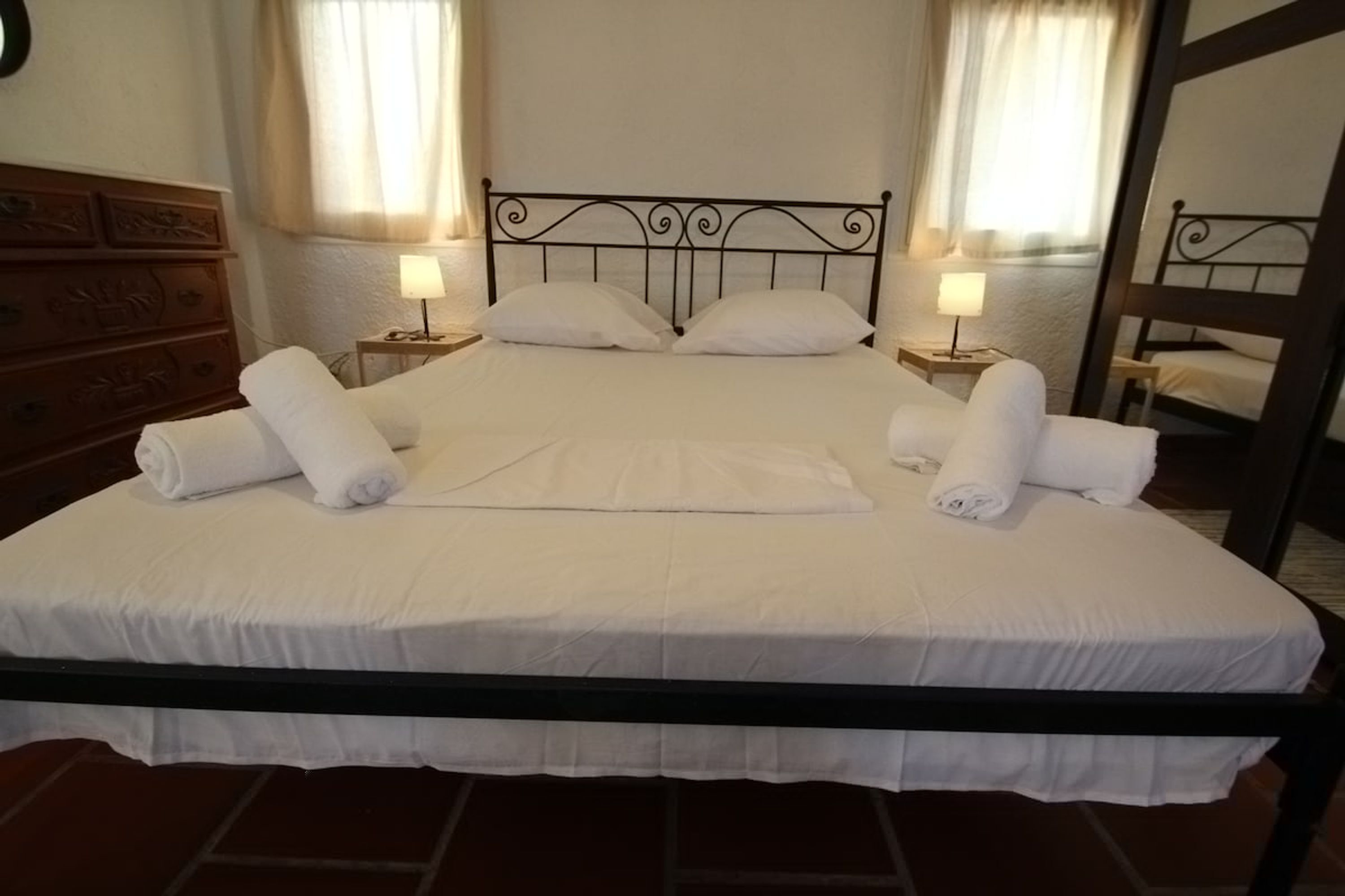 Cama bem arrumada com lençóis brancos, duas mesas de cabeceira com abajures e um aparador de madeira.