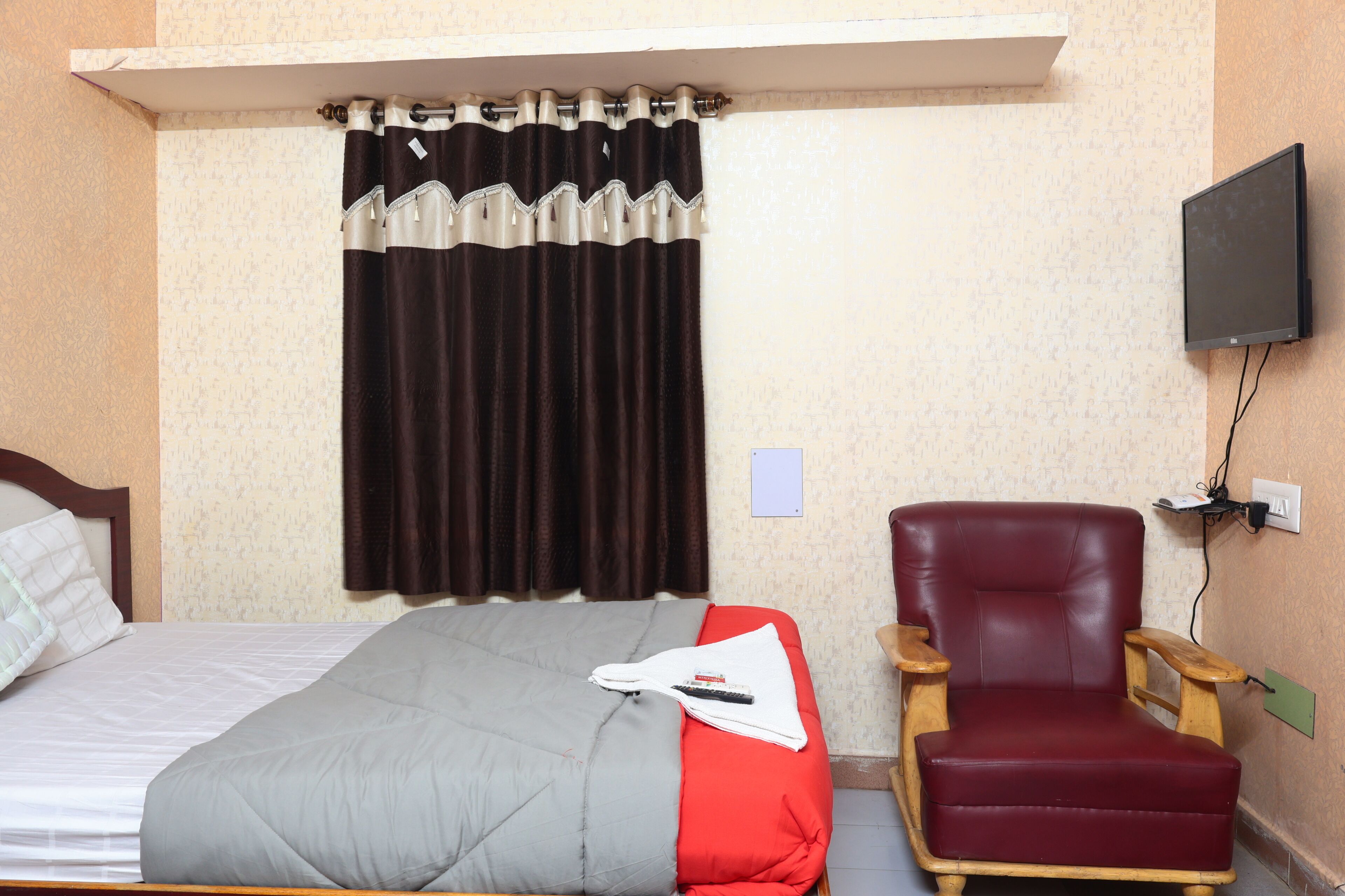 Quarto de hotel com uma cama, uma poltrona vermelha, uma televisão de tela plana e uma janela com cortinas.