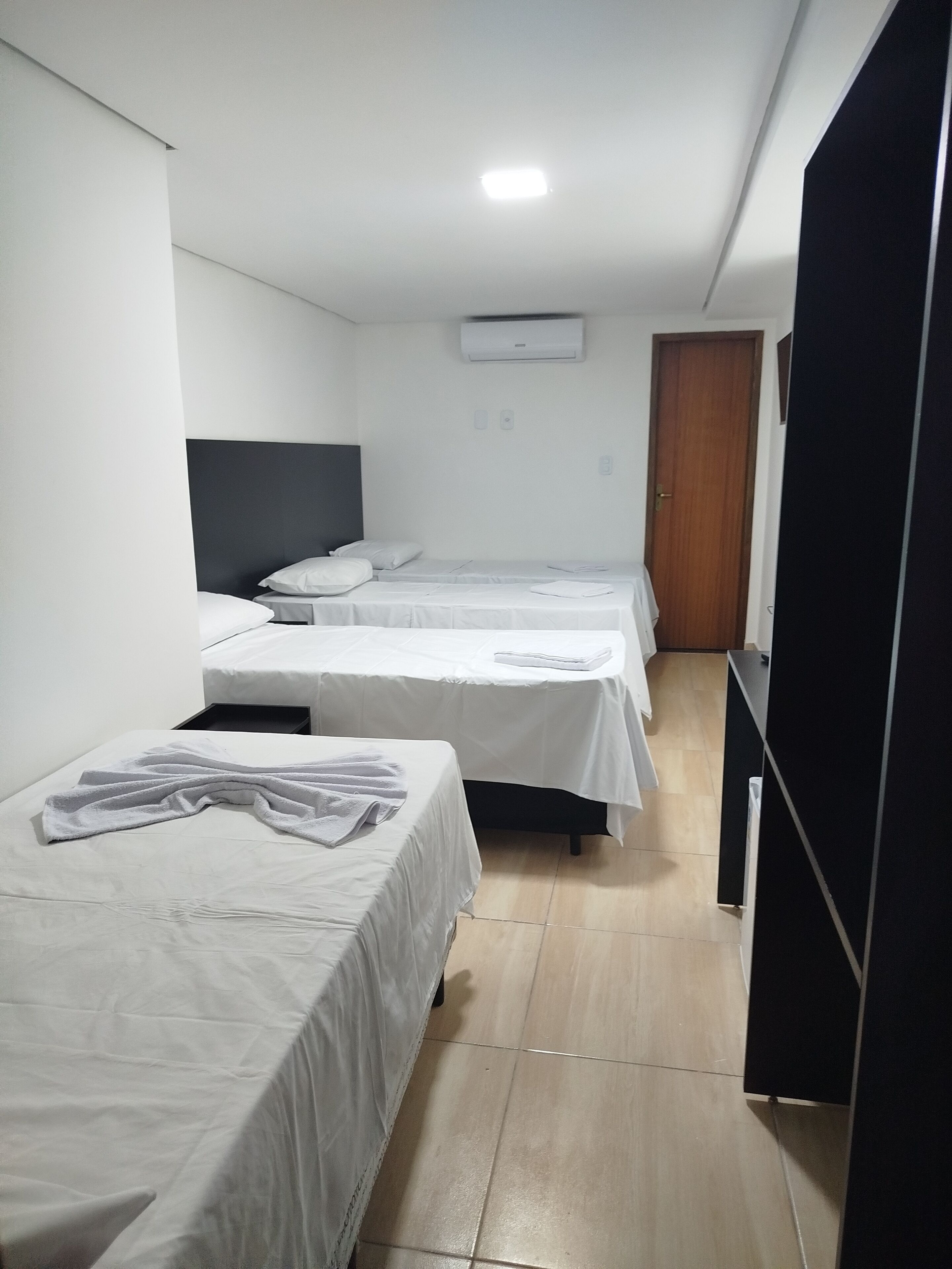 Quarto de hotel com três camas individuais, uma porta de madeira e um guarda-roupa preto.