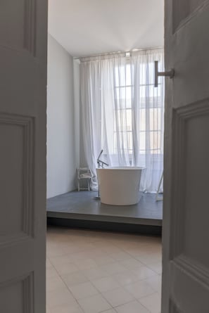 Casa de banho com banheira, um banquinho pequeno e janela com cortinas leves.