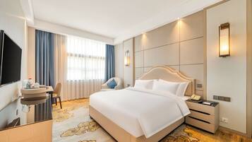 Quarto de hotel com uma cama grande, mesas de cabeceira, escrivaninha com cadeira, televisão de tela plana e uma grande janela com cortinas.