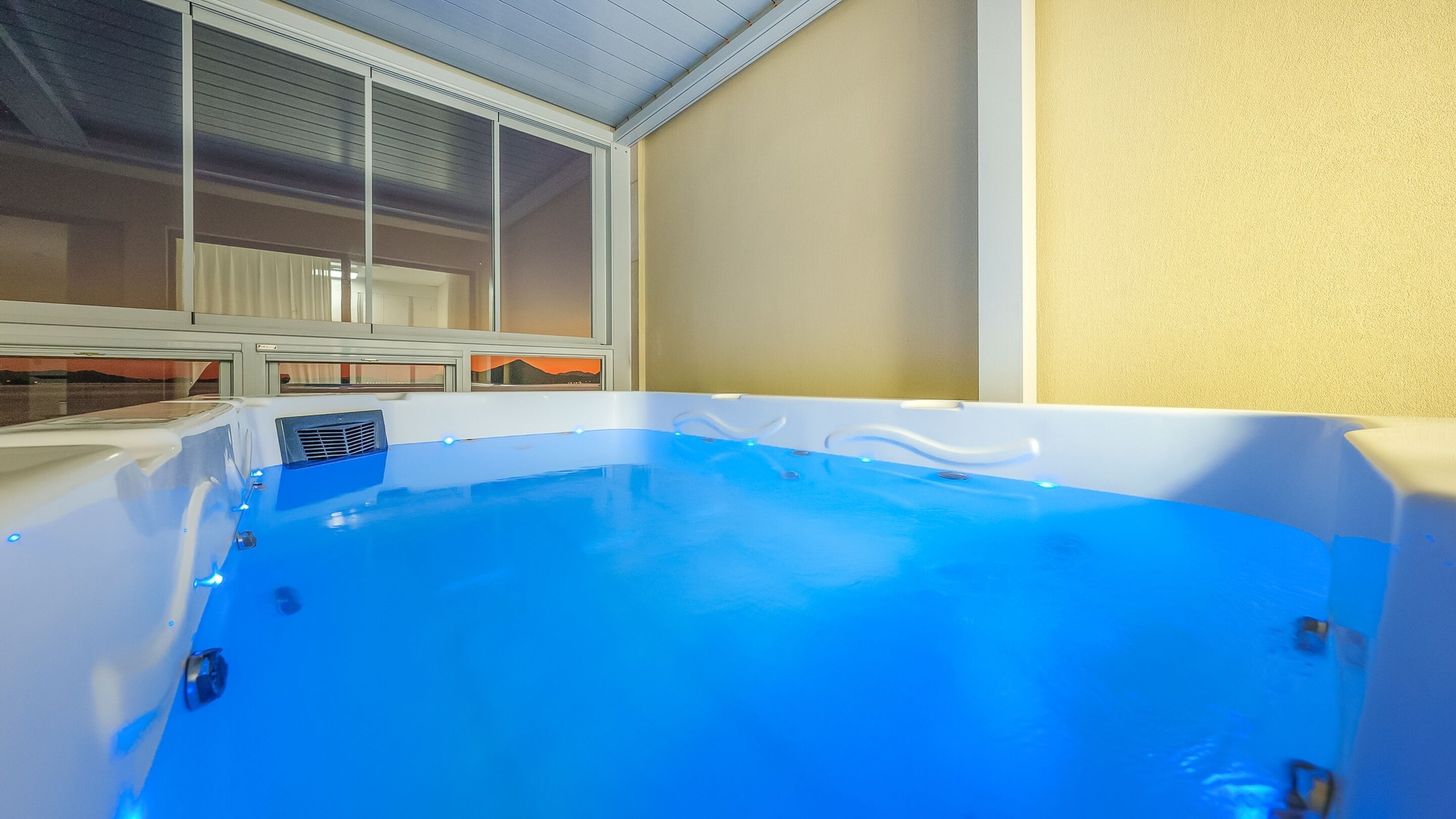 Uma banheira de hidromassagem com interior azul transparente, cercada por uma janela e uma parede bege.