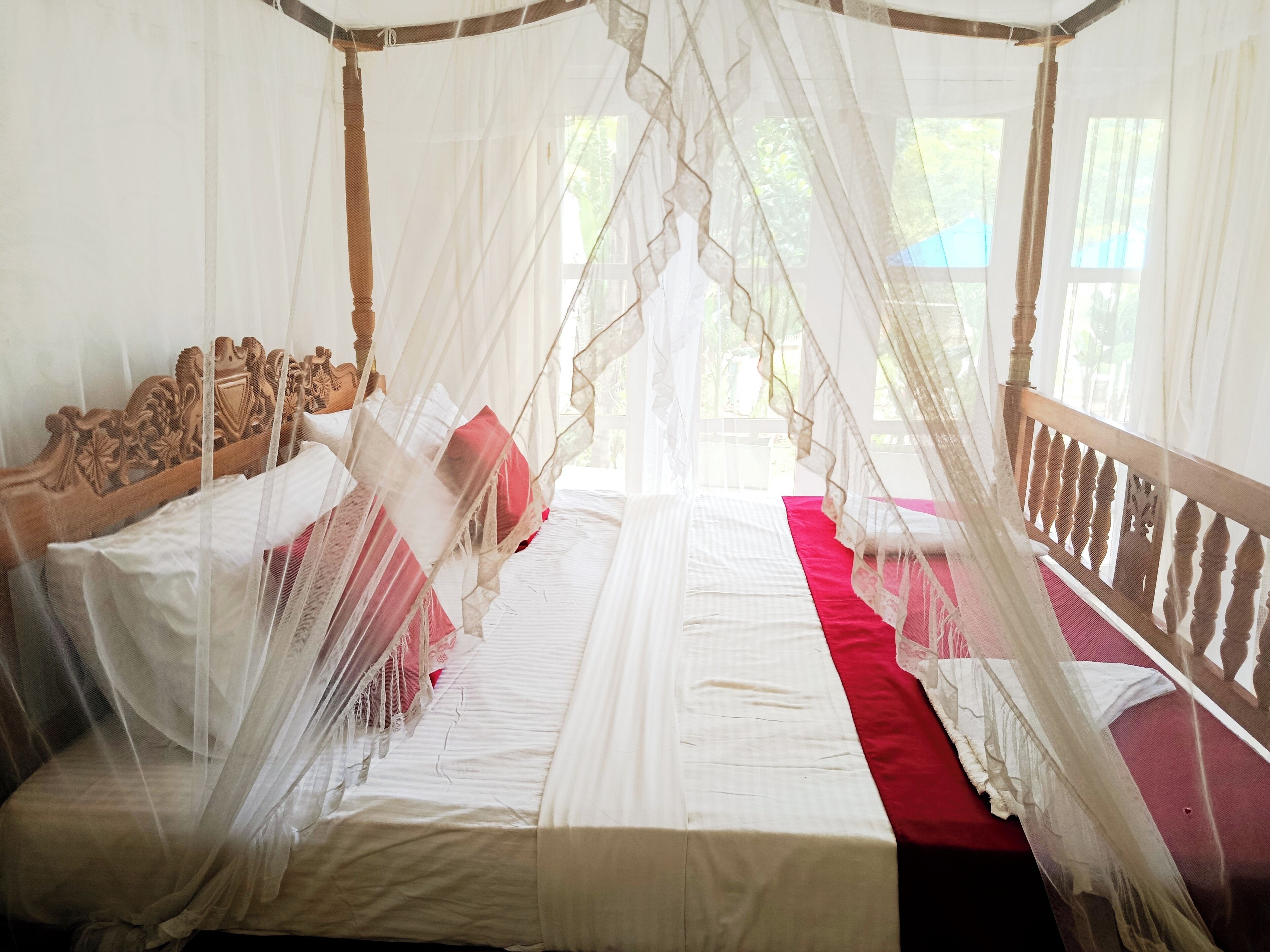Cama com dossel e cortinas, roupa de cama branca com detalhes vermelhos, cabeceira e peseira de madeira, e vista para o verde lá fora.