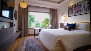 Quarto de hotel com uma cama grande, televisão, janela com cortinas, uma mesinha e uma cadeira.