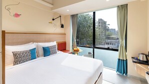 Quarto de hotel com uma cama grande, uma mesa pequena com uma cesta de frutas, uma escrivaninha com uma chaleira e vista para a cidade através de uma janela ampla.
