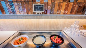 Uma área de buffet com potes de frutas cortadas e coalhada, uma lousa e um espaço com copos e taças.