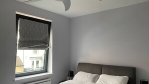 Quarto com cama, janela com persiana de padrão geométrico, ventilador de teto e radiador.