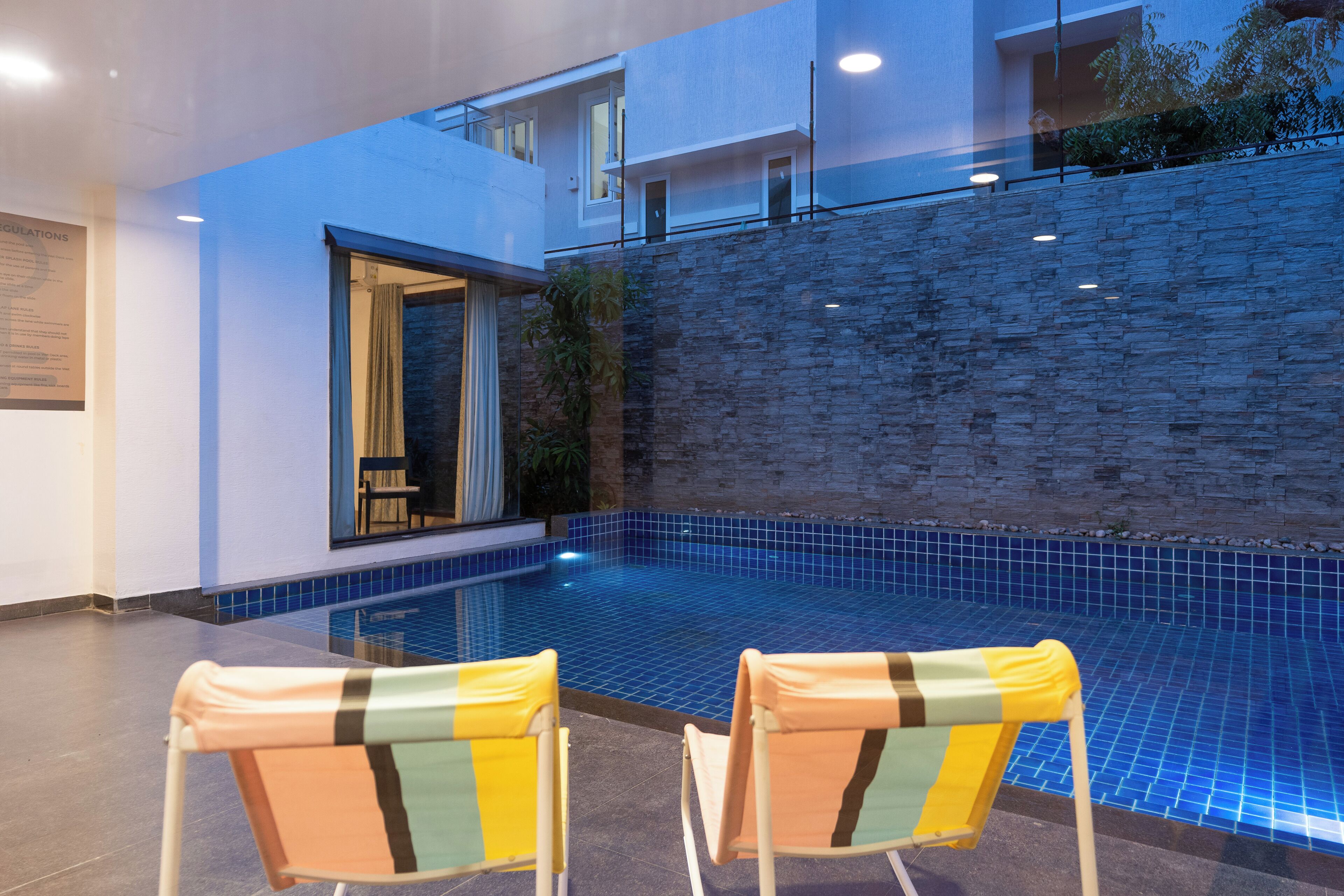 Uma área interna de piscina moderna com duas espreguiçadeiras, uma parede de pedra e uma porta de vidro que dá para um quarto.