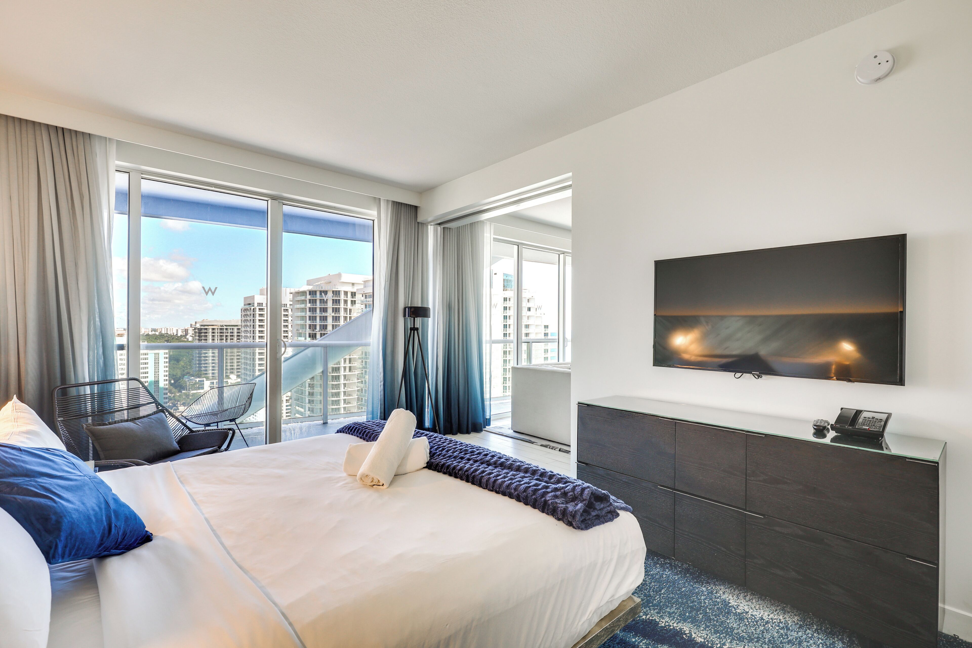 Quarto de hotel moderno com uma cama grande, uma escrivaninha, uma cadeira, uma televisão e uma varanda com vista para a cidade.