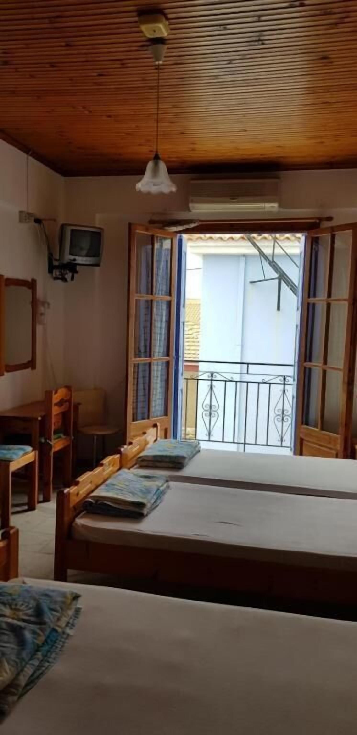 Quarto com teto de madeira, cama com colchão branco, escrivaninha de madeira, uma cadeira e televisão na parede.