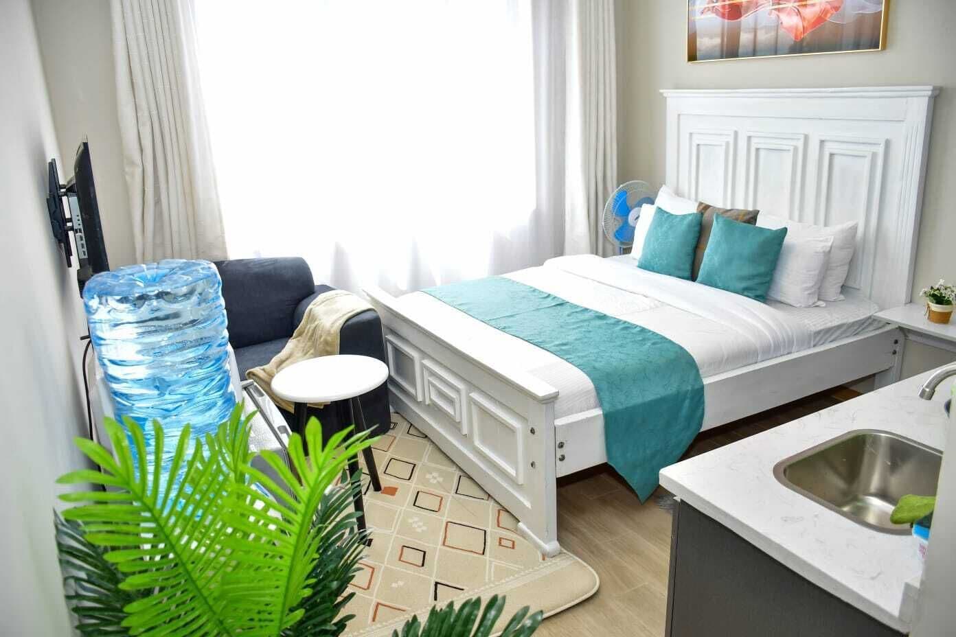 Quarto de hotel com uma cama, uma pequena mesa, uma cadeira, um dispensador de água, uma pia e um vaso de plantas.