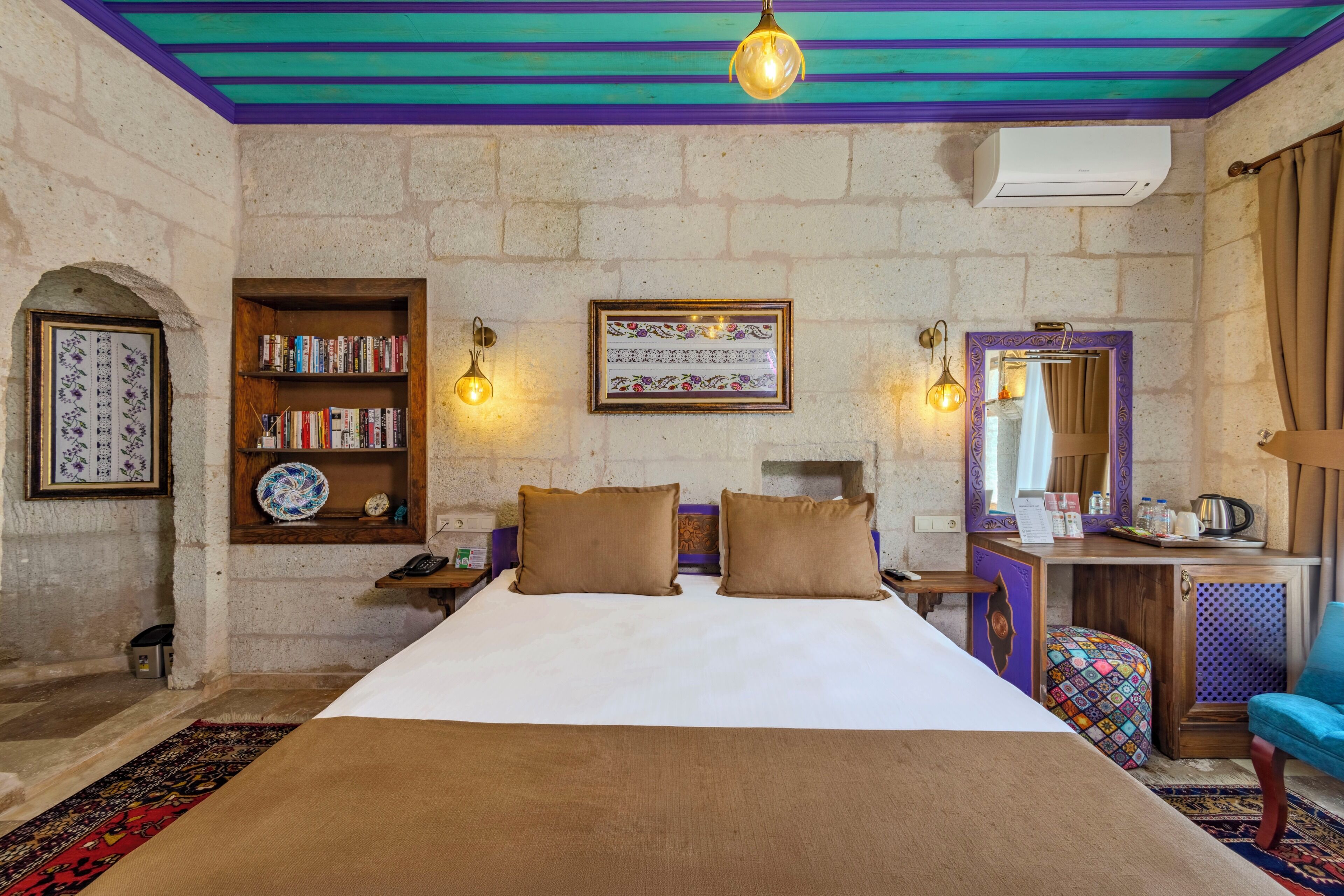 Quarto de hotel com cama grande, estante de livros, espelho decorativo e mesa com chaleira.