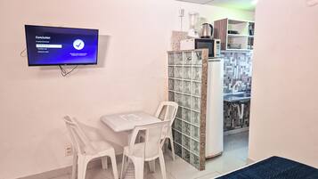 Um quarto pequeno com uma TV exibindo uma lista de canais, uma mesa branca com duas cadeiras, uma área de cozinha com micro-ondas e liquidificador, e um tapete azul com texto no chão.