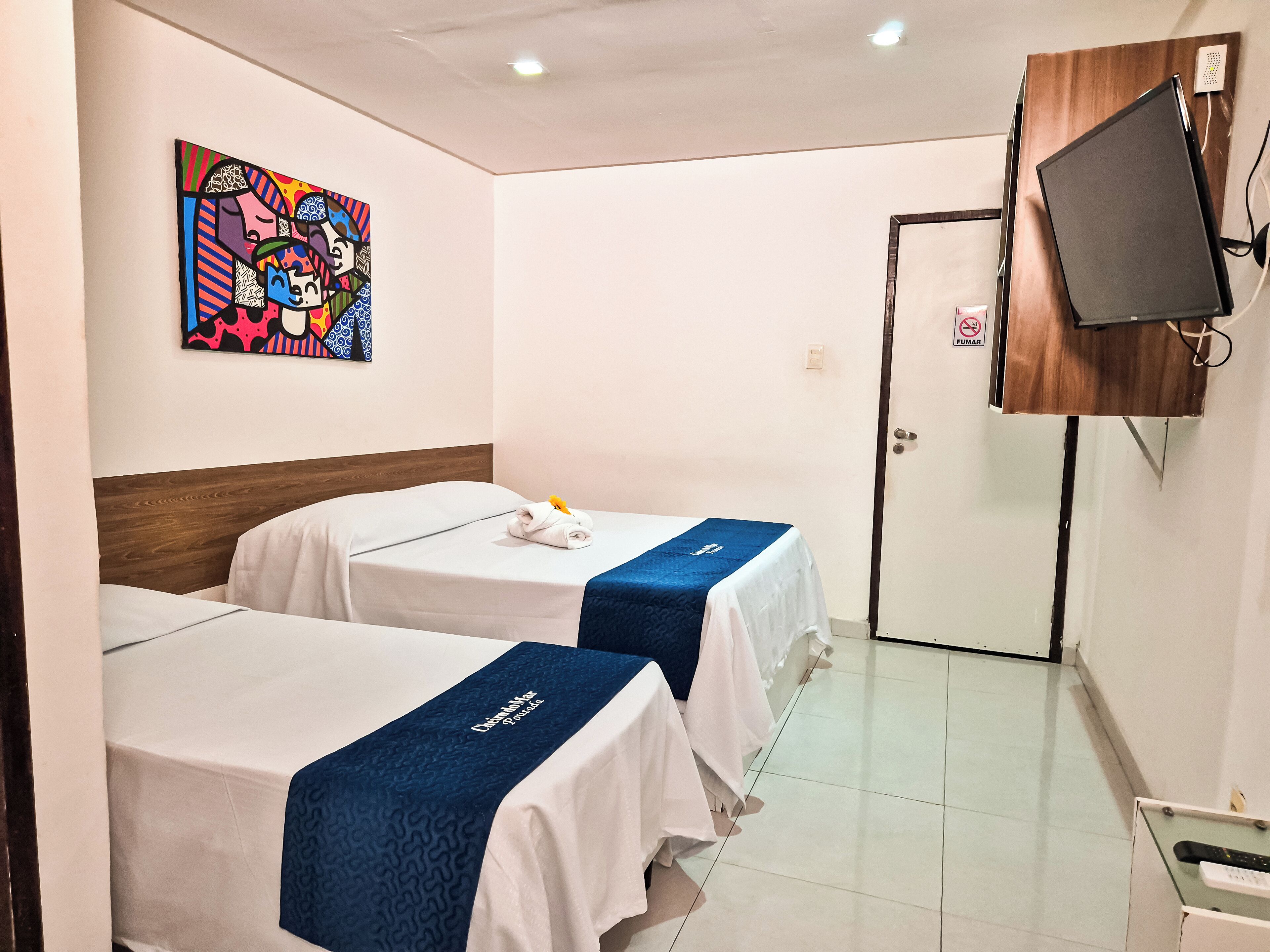 Quarto de hotel com duas camas, uma televisão fixada na parede e uma obra de arte colorida acima das camas.