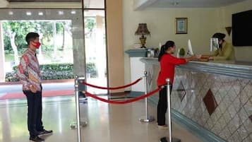 Uma área de recepção com recepcionista, um hóspede aguardando e uma barreira de corda vermelha.