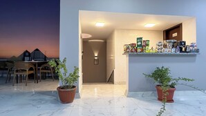 Um saguão de hotel moderno, com piso de mármore, plantas em vasos, balcão de recepção e exposição de bebidas engarrafadas.