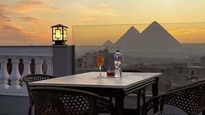 Mesa com duas cadeiras de vime, um copo de suco de laranja, uma garrafa de água e um cinzeiro vermelho numa varanda com vista para uma paisagem urbana com pirâmides ao fundo.