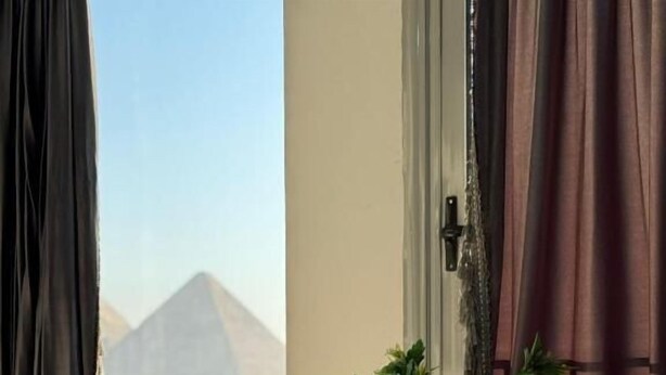 Quarto de hotel com uma cama, janela com cortinas, vista de uma pirâmide e banheiro com chuveiro.