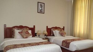 Quarto de hotel com duas camas de solteiro, cada uma com cabeceira de madeira, roupa de cama branca e almofadas decorativas. Há uma mesa de cabeceira com uma luminária entre as camas, e um quadro emoldurado na parede.