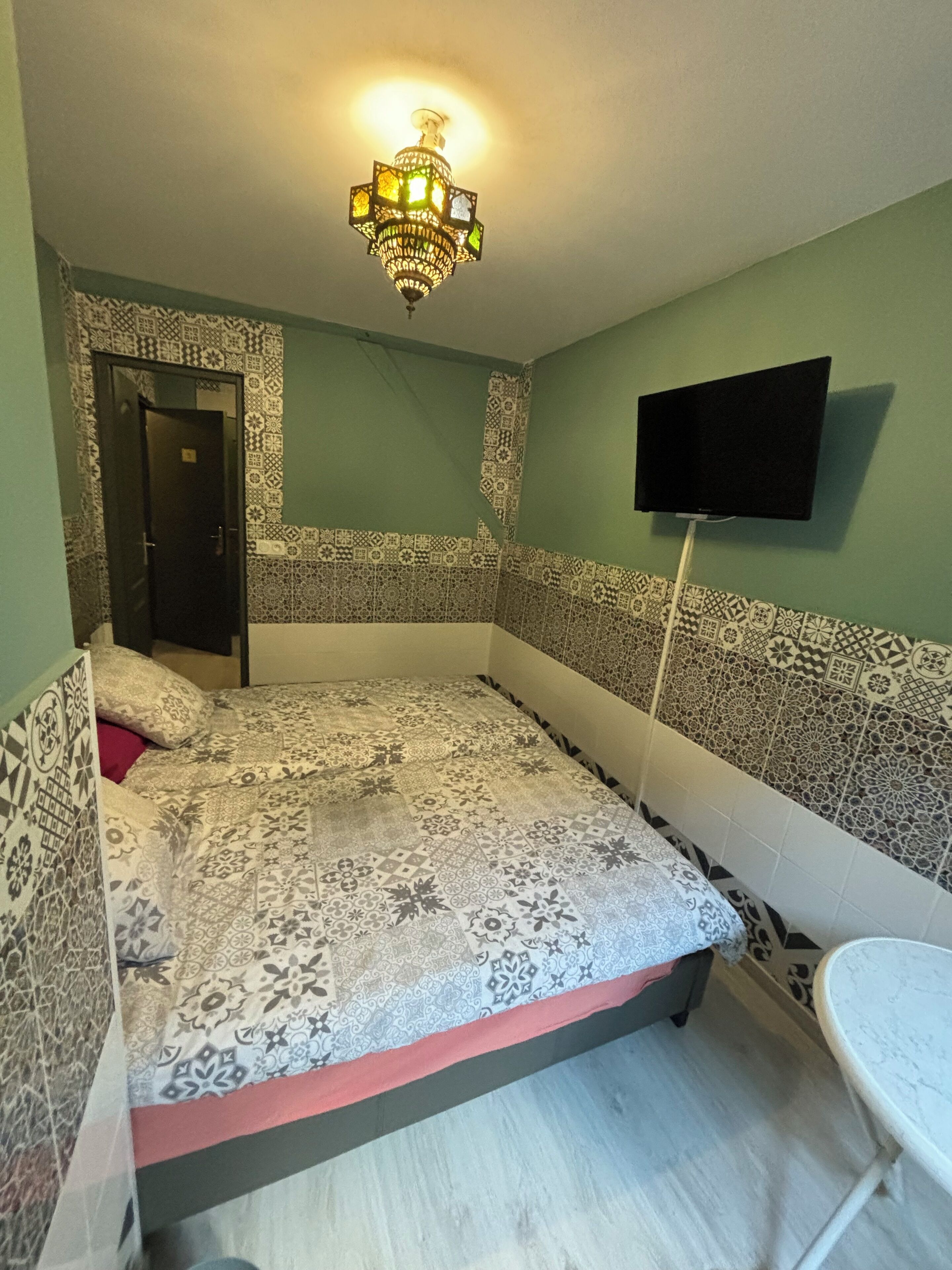 Quarto com paredes estampadas, cama com colcha florida, televisão na parede e um lustre.