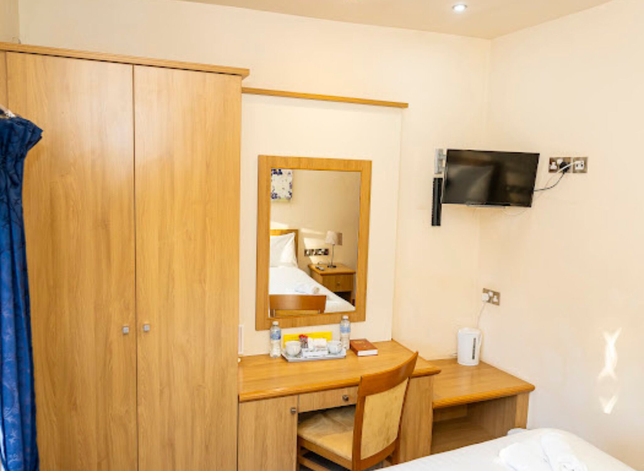 Quarto de hotel com guarda-roupa de madeira, secretária com cadeira, cama, televisão fixada na parede e espelho sobre a secretária.
