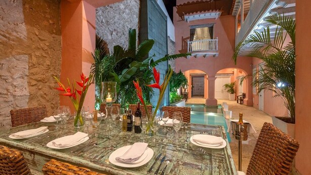 Uma área de refeições com mesa de vidro posta para quatro pessoas, rodeada por cadeiras de vime, com uma piscina e um edifício ao fundo.