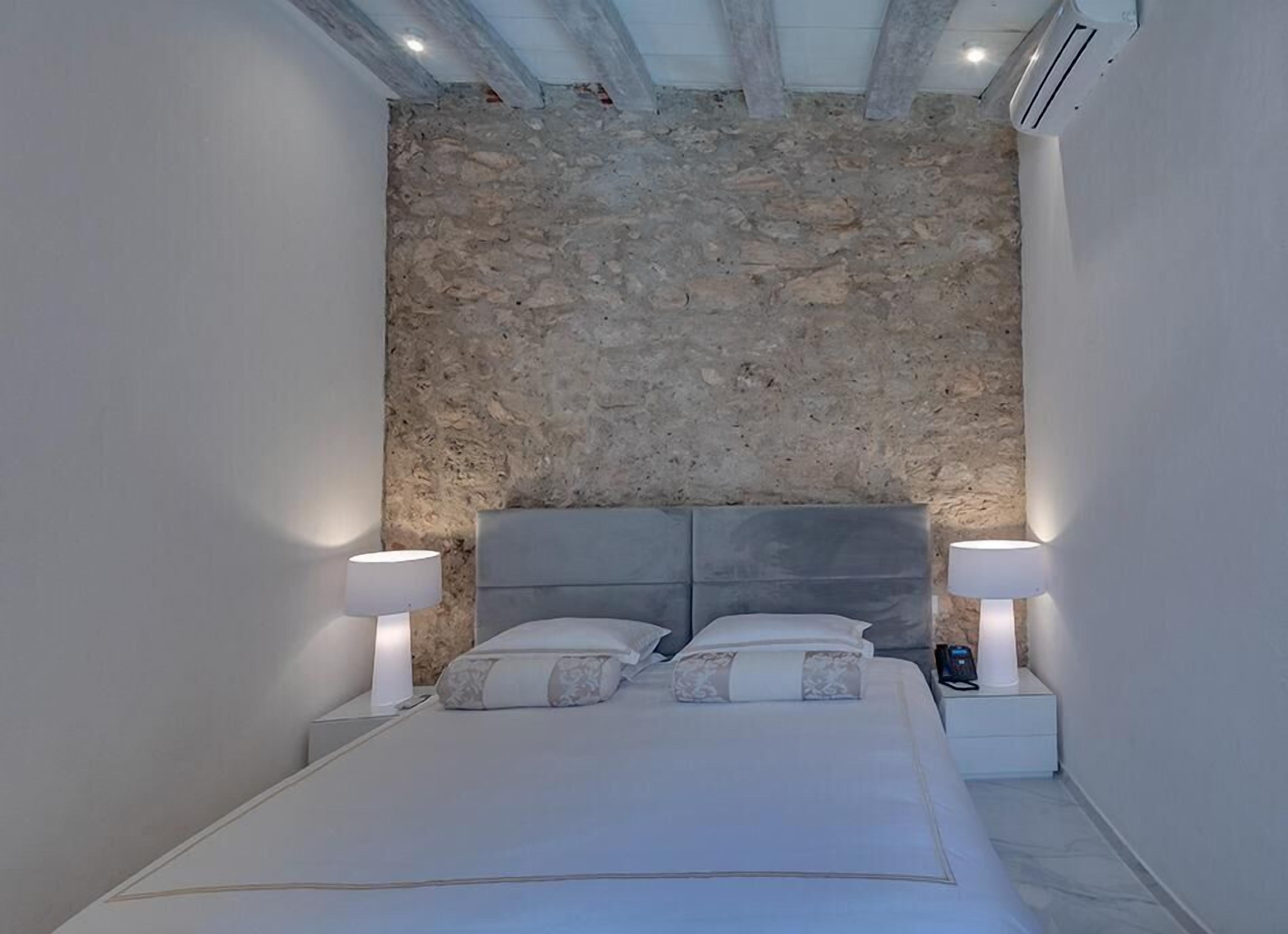 Quarto com parede de pedra, cama com duas almofadas e duas luminárias de cabeceira.