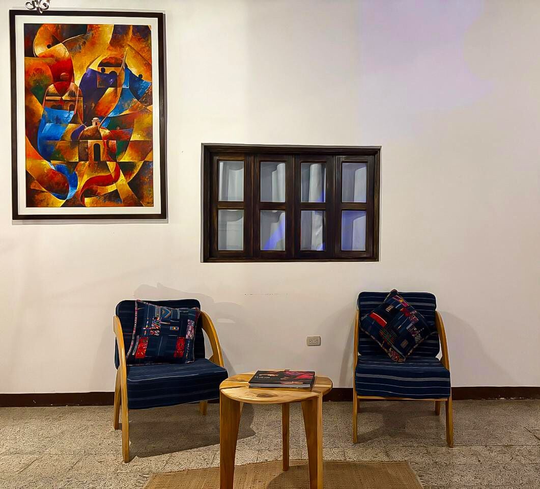 Quarto com duas cadeiras, uma mesinha, uma janela e um quadro abstrato emoldurado.