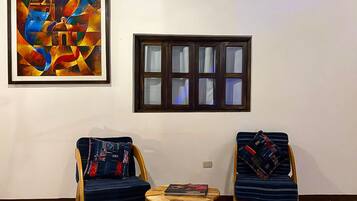 Quarto com duas cadeiras, uma mesinha, uma janela e um quadro abstrato emoldurado.