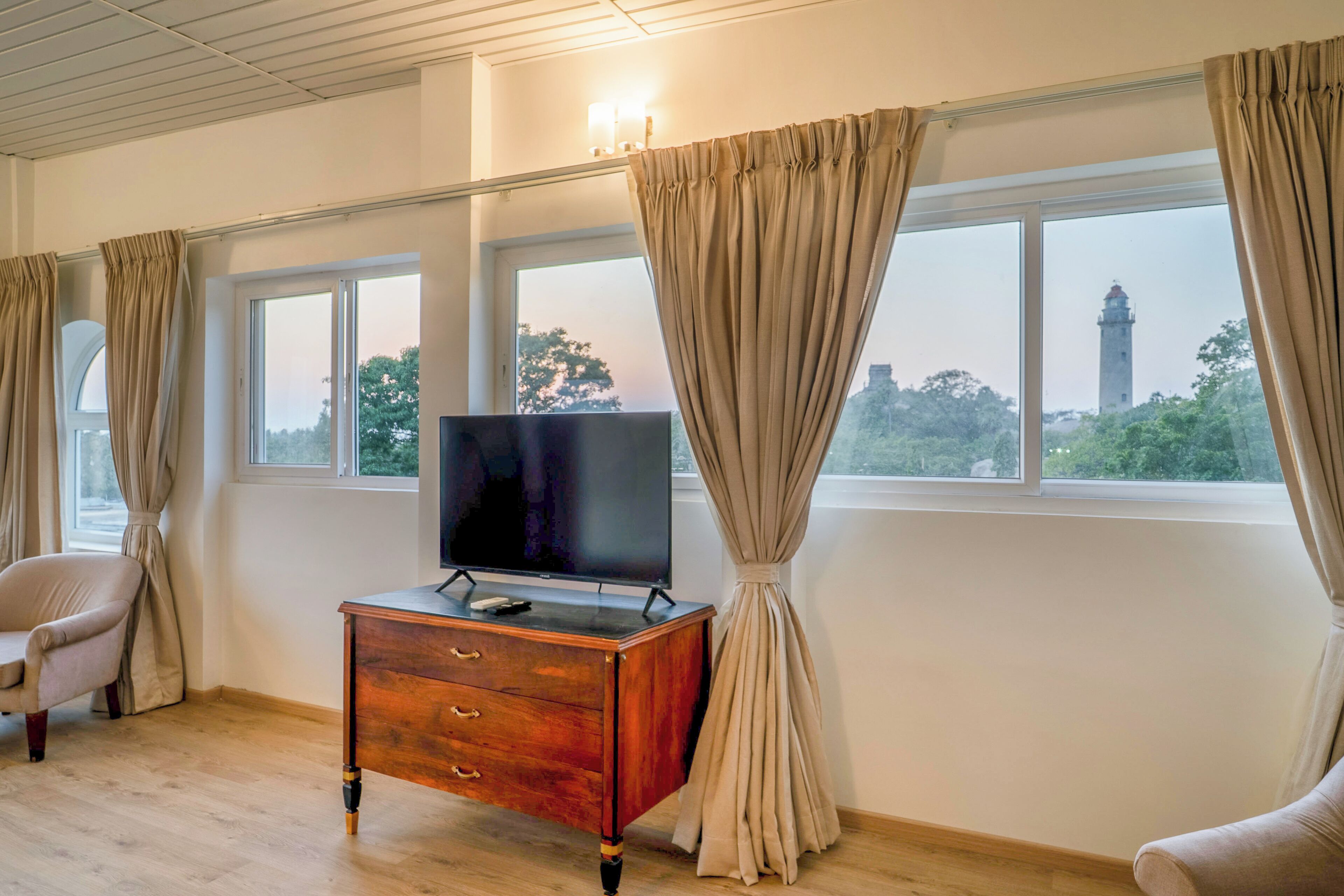 Quarto com cômoda de madeira, televisão e uma grande janela com cortinas.