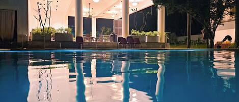 Piscina com área de terraço com cadeiras e mesas, cercada por colunas e plantas.