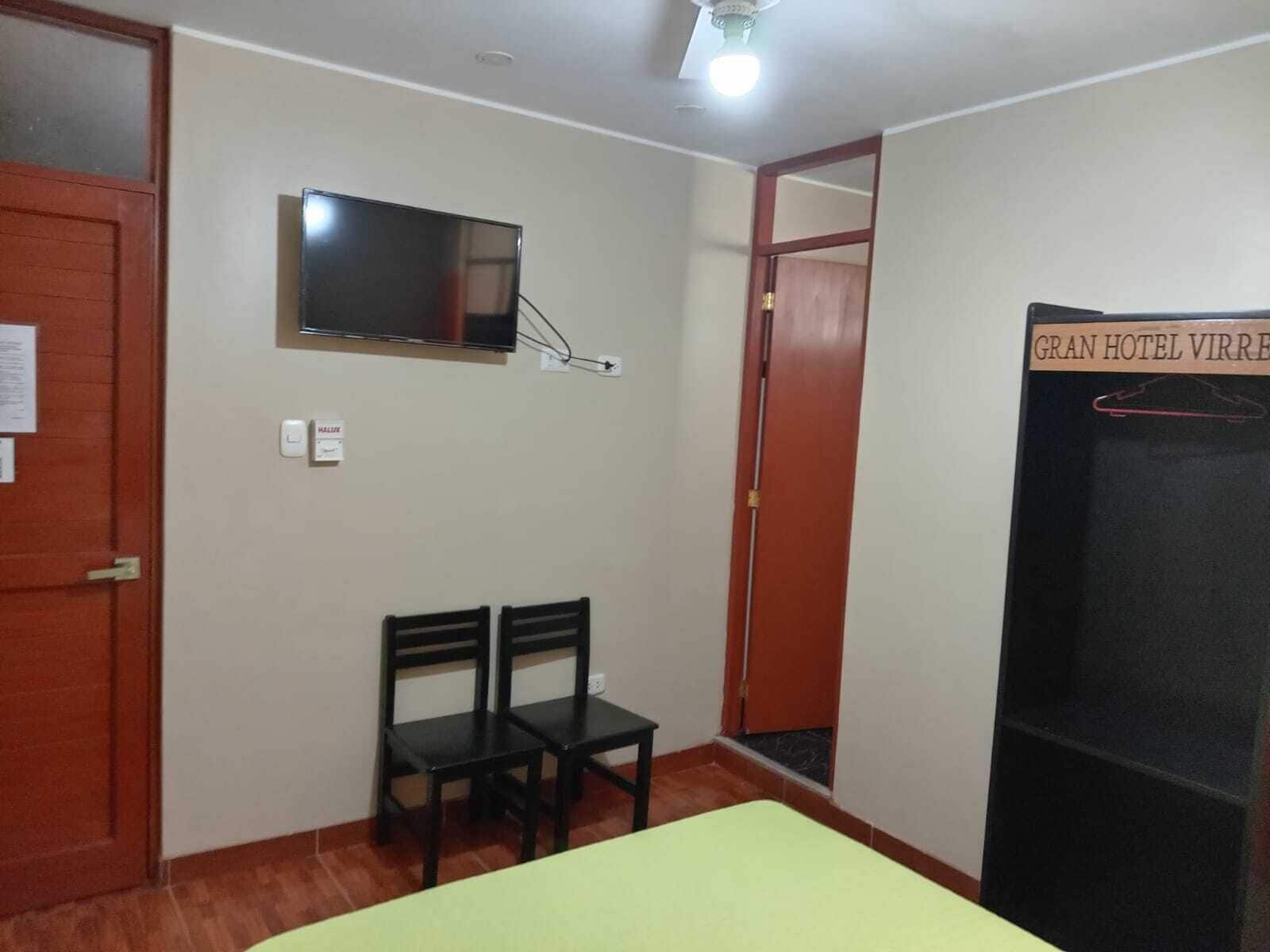 Quarto de hotel com cama, televisão fixada na parede, duas cadeiras e porta com a inscrição "GRAN HOTEL VIRRE".