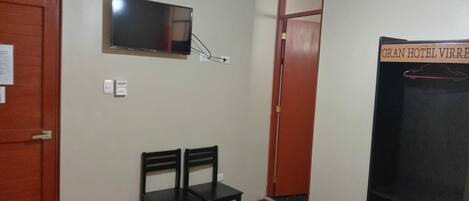 Quarto de hotel com cama, televisão fixada na parede, duas cadeiras e porta com a inscrição "GRAN HOTEL VIRRE".