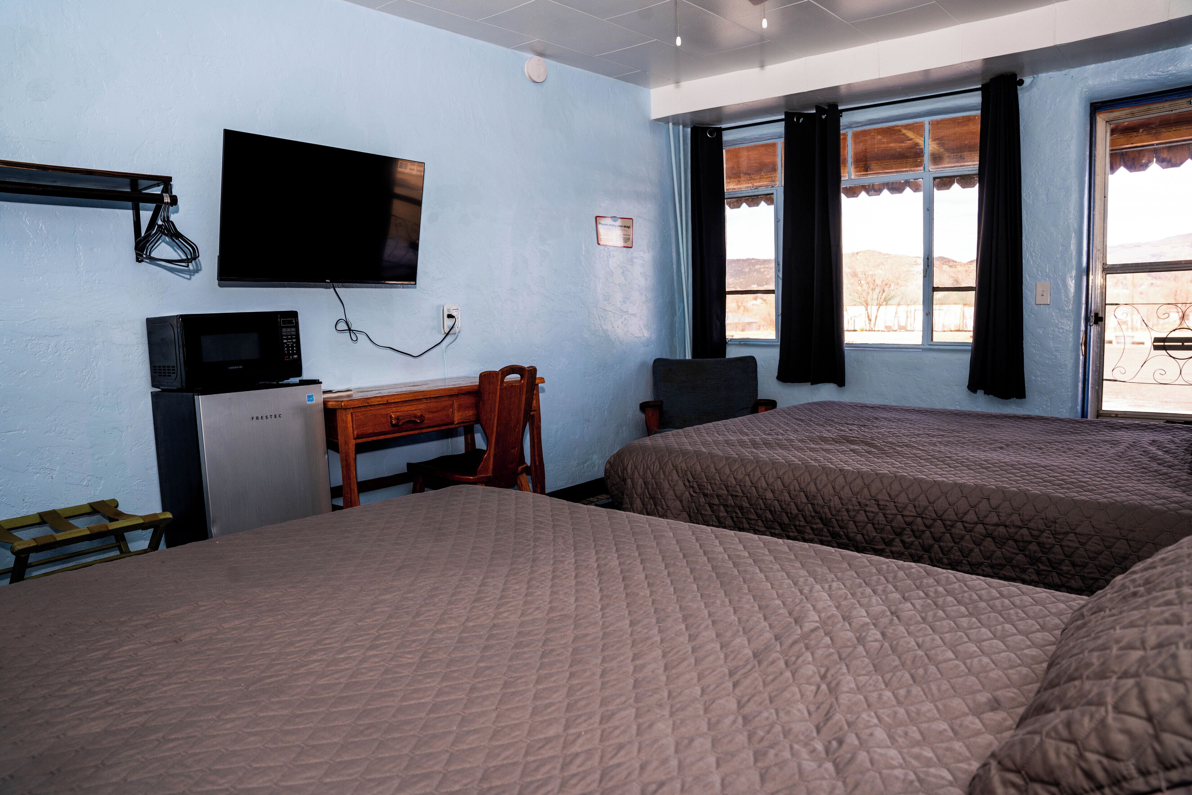 Quarto com duas camas, uma escrivaninha, uma cadeira, uma televisão e uma janela com cortinas.