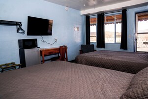 Quarto com duas camas, uma escrivaninha, uma cadeira, uma televisão e uma janela com cortinas.
