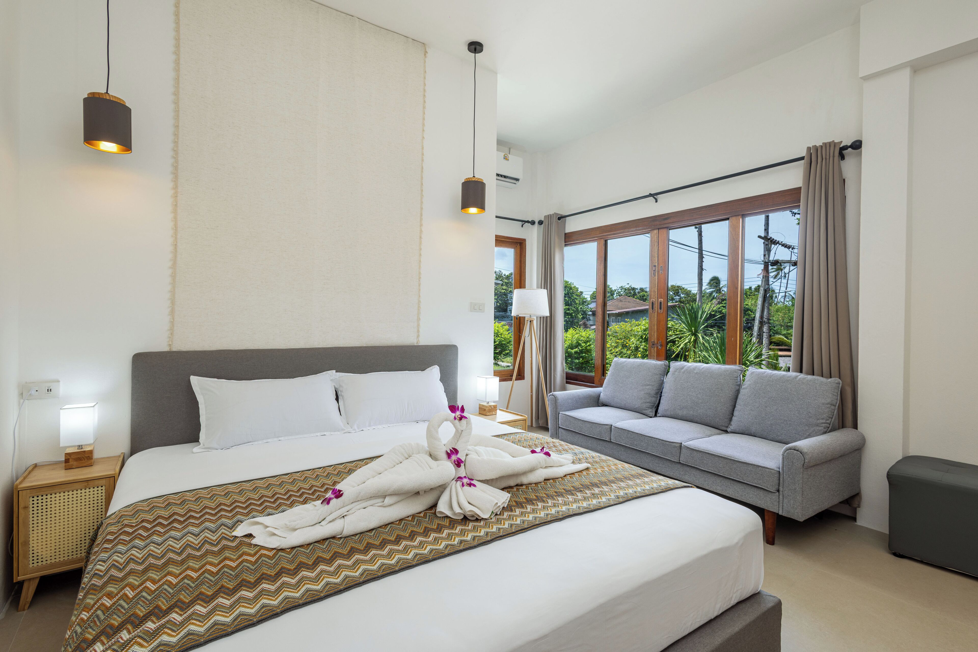 Quarto de hotel com uma cama grande, um sofá cinzento, uma janela com cortinas e um ar condicionado fixado na parede.
