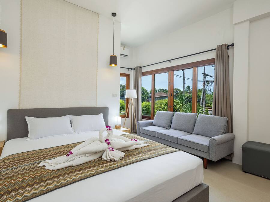 Quarto de hotel com uma cama grande, um sofá cinzento, uma janela com cortinas e um ar condicionado fixado na parede.