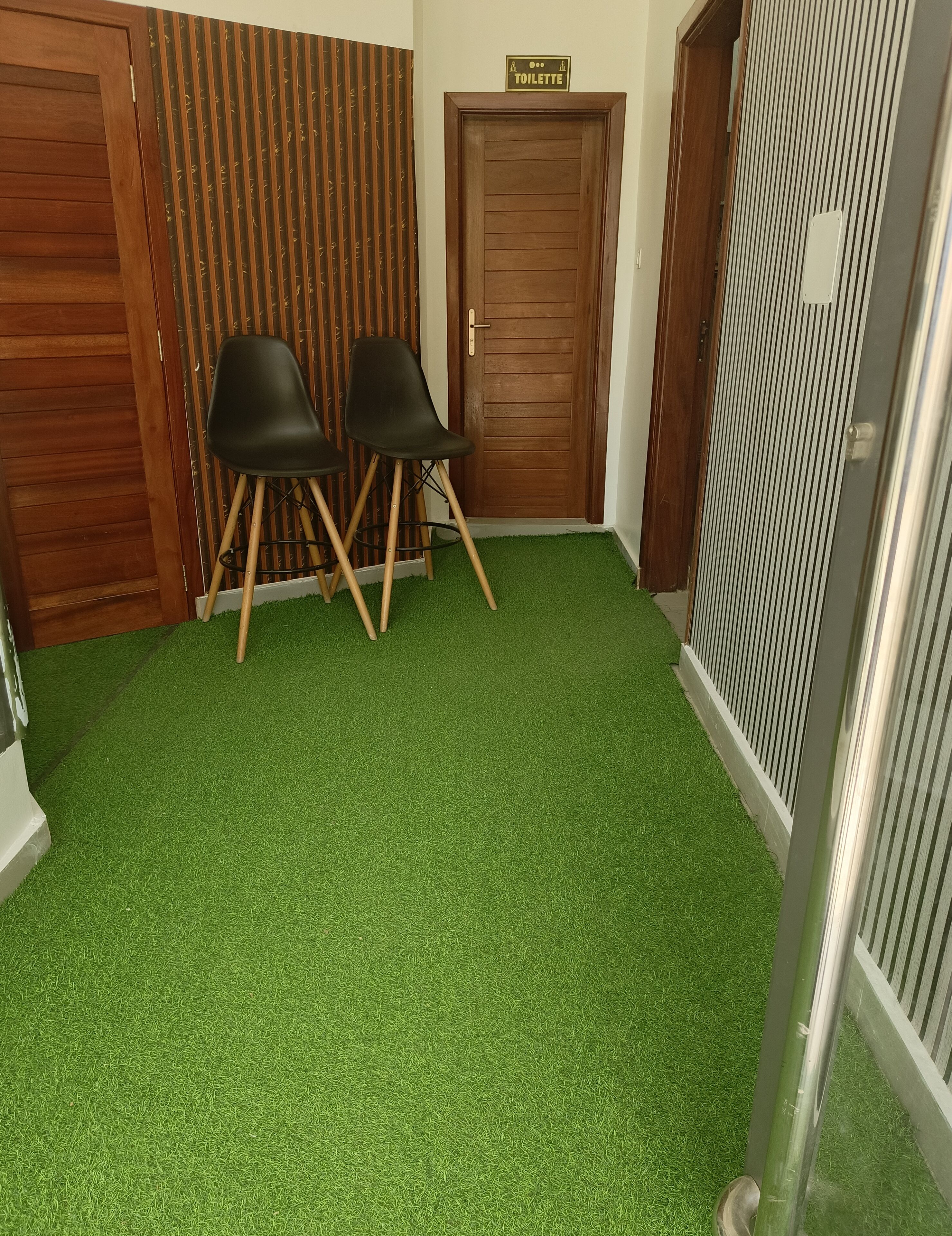 Quarto com carpete verde artificial, duas cadeiras pretas, porta de madeira e um aviso indicando um banheiro.