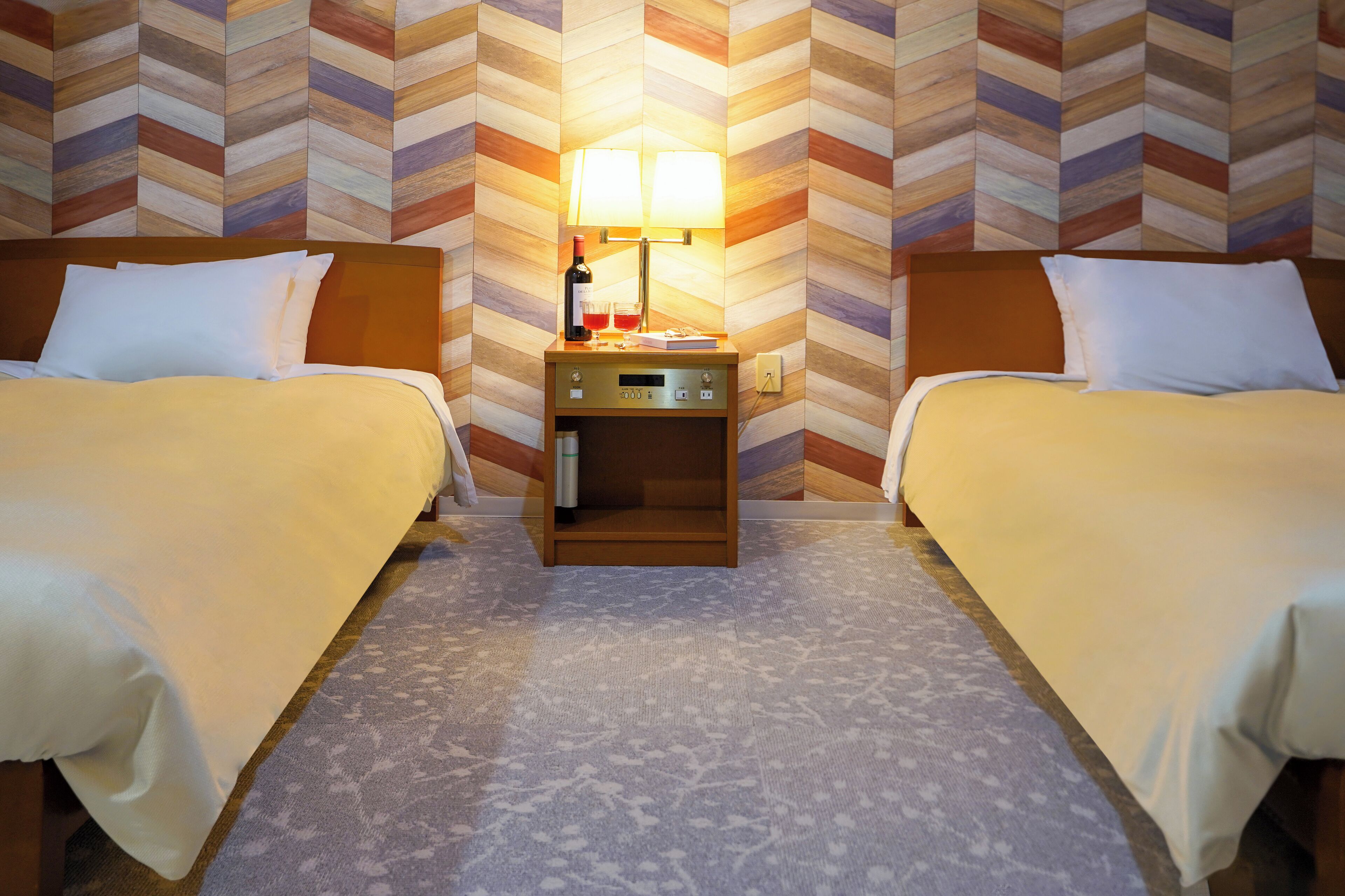 Quarto de hotel com duas camas, uma mesa de cabeceira com abajur e parede estampada.