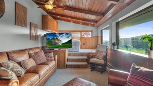 Uma sala de estar acolhedora com um sofá de couro, uma televisão e vista para uma paisagem montanhosa através de uma grande janela.