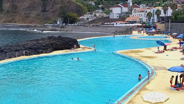 Uma grande piscina ao ar livre com água azul cristalina, rodeada por uma praia de areia e um litoral rochoso.