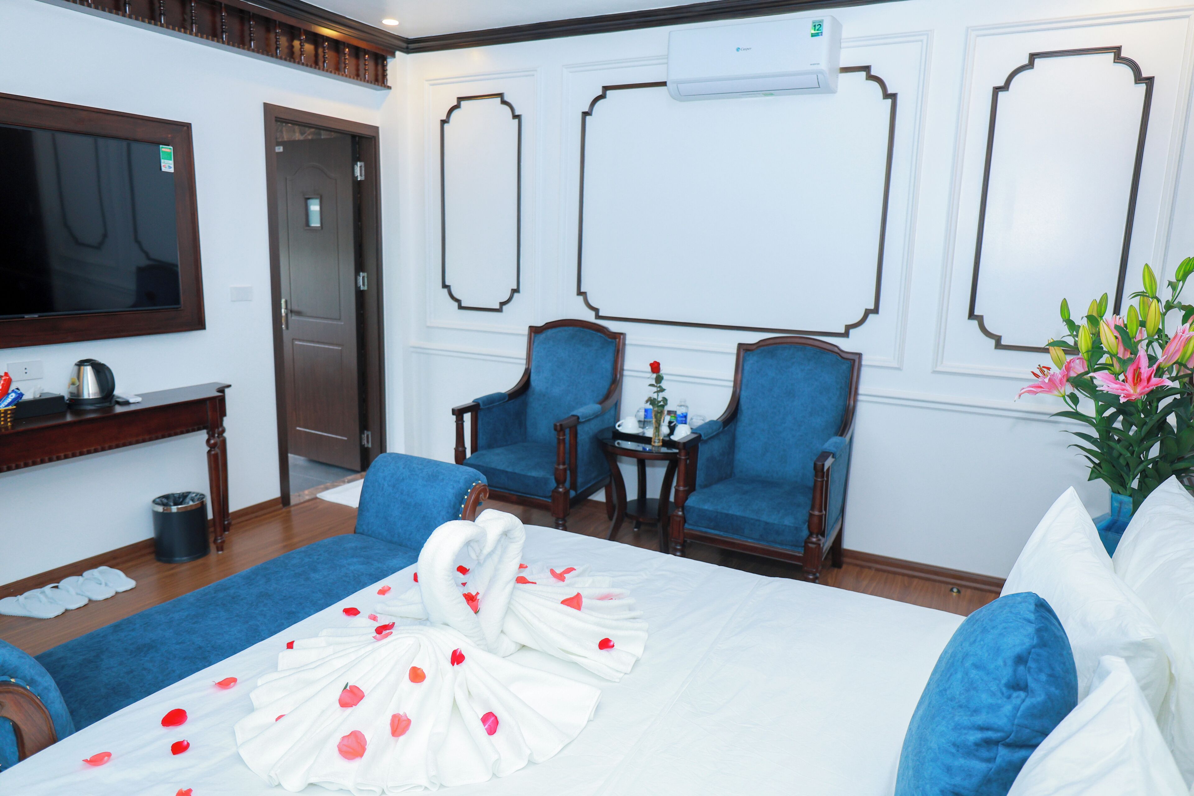 Quarto de hotel com uma cama, duas poltronas azuis, uma mesinha, um espelho e um vaso de planta.