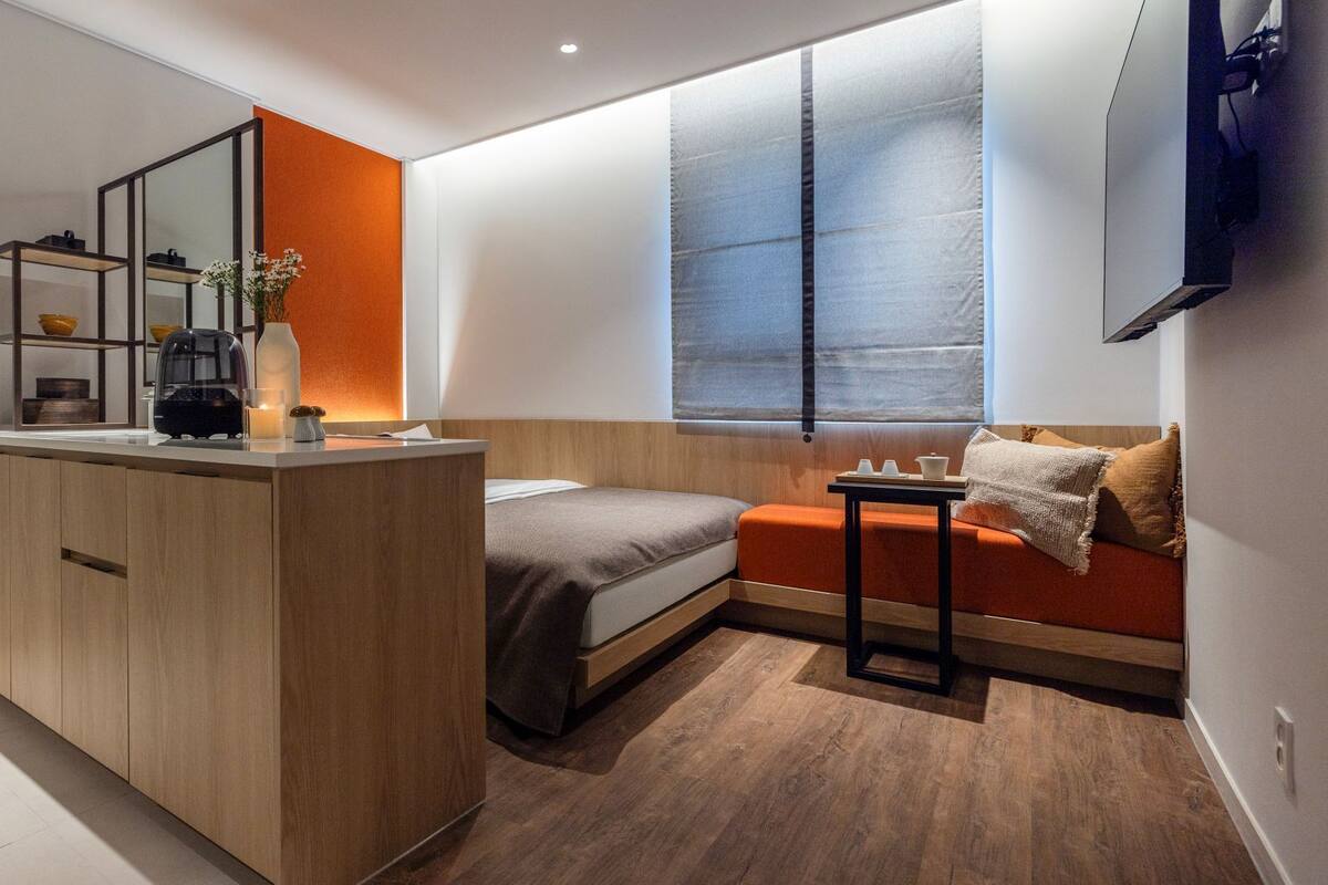 Quarto de hotel moderno com um armário de madeira, uma cama com cabeceira laranja, uma mesa pequena e uma janela grande com cortinas.