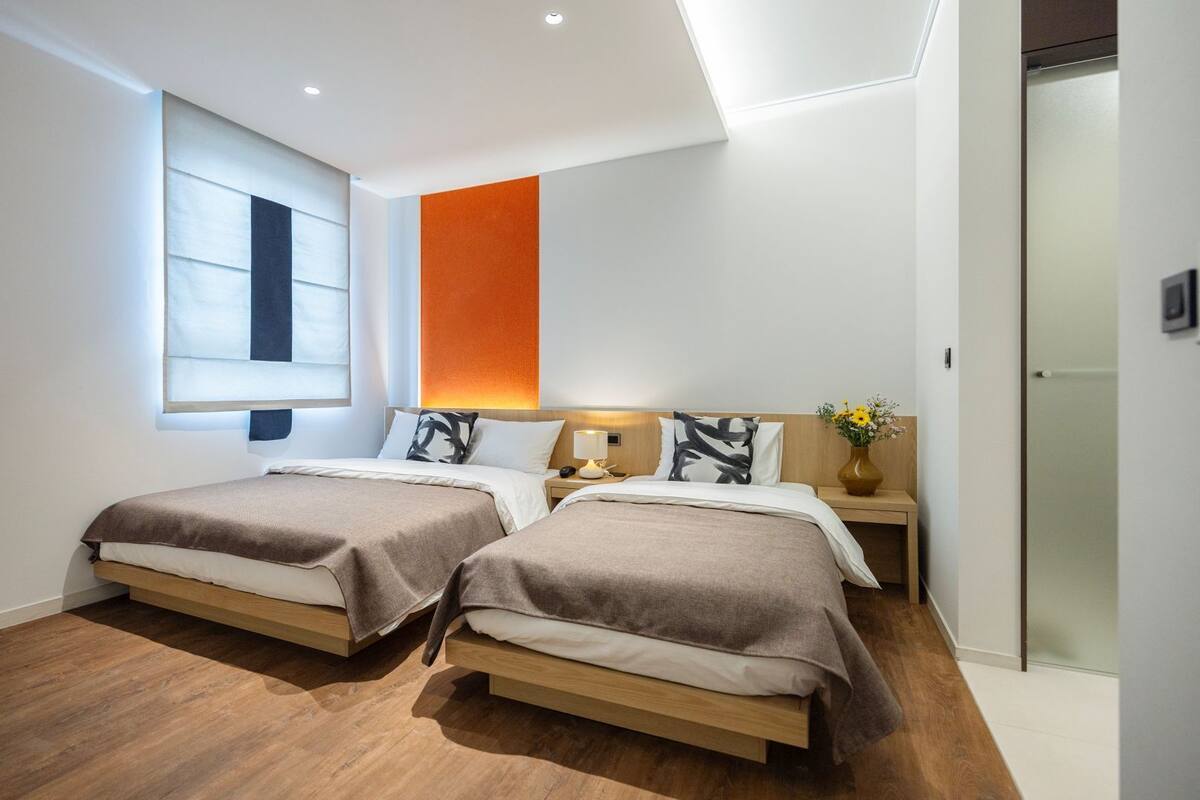 Quarto de hotel moderno com duas camas, uma janela grande, uma luminária fixada na parede e piso de madeira.