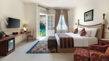 Quarto de hotel com cama, mesa de trabalho com televisão, cadeira, mesinha, mesa de cabeceira e janela com cortinas.