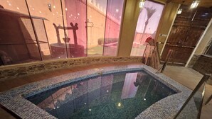 Piscina com bordas de azulejos mosaicados, uma parede de pedra e vista para um edifício com janelas em arco.